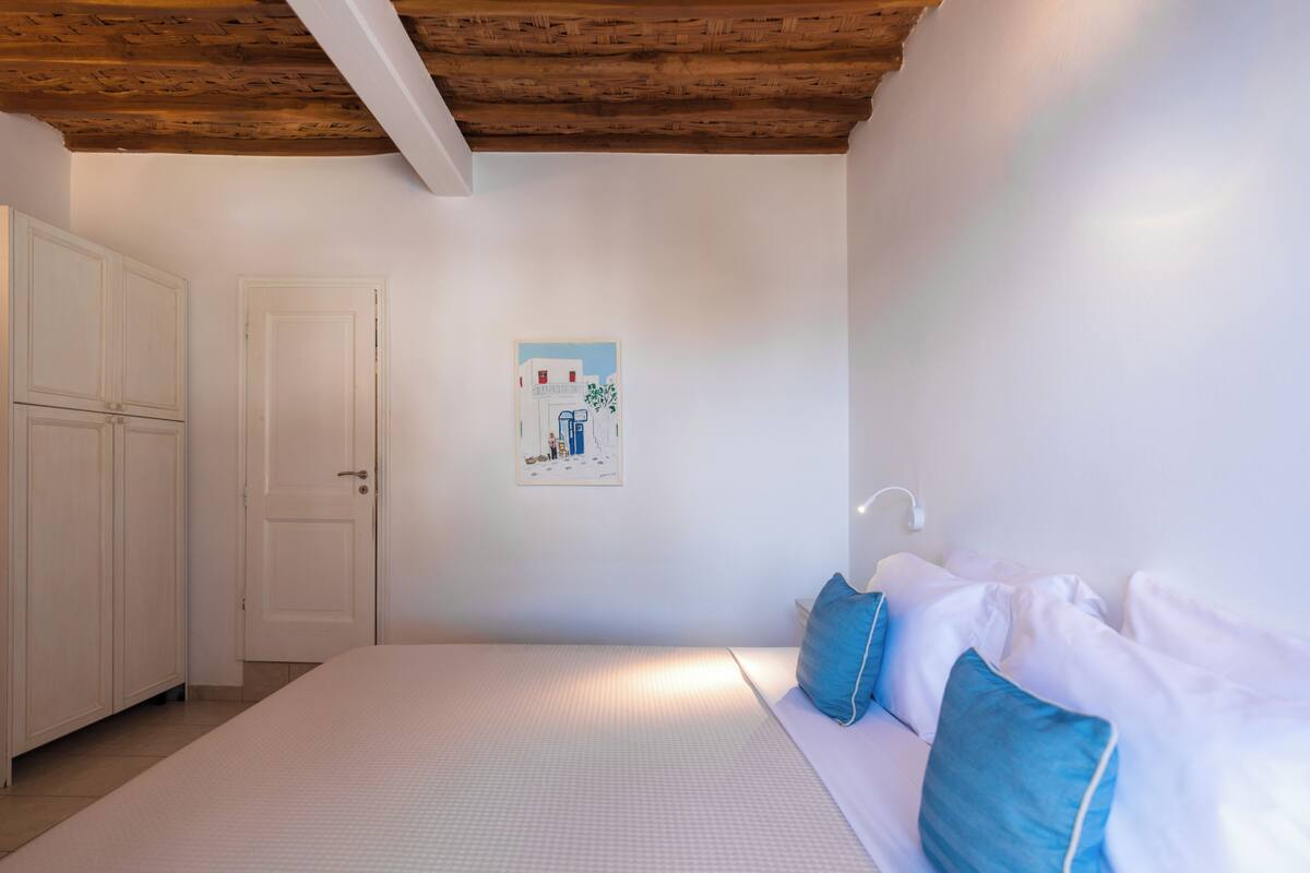 Quarto com cama, travesseiros, porta, parede com quadro e teto de madeira.