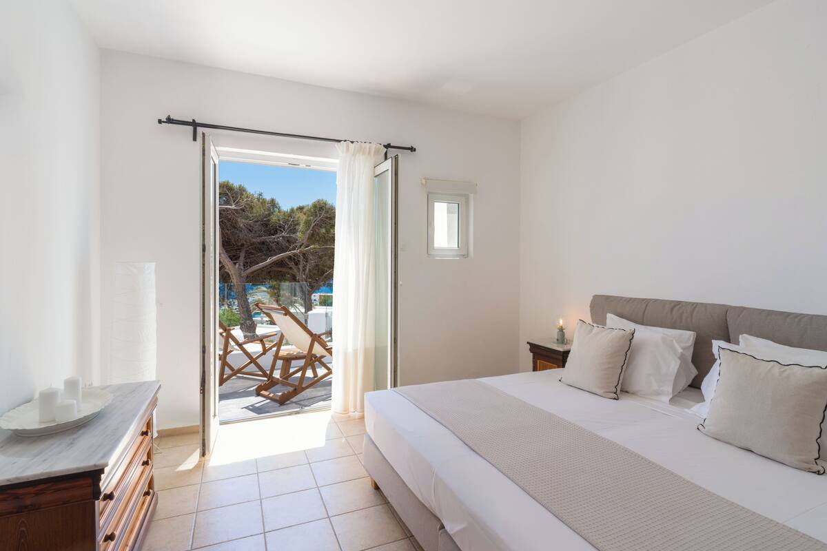 Quarto com cama, mesa de cabeceira, cômoda, janela com cortinas e vista para o exterior.