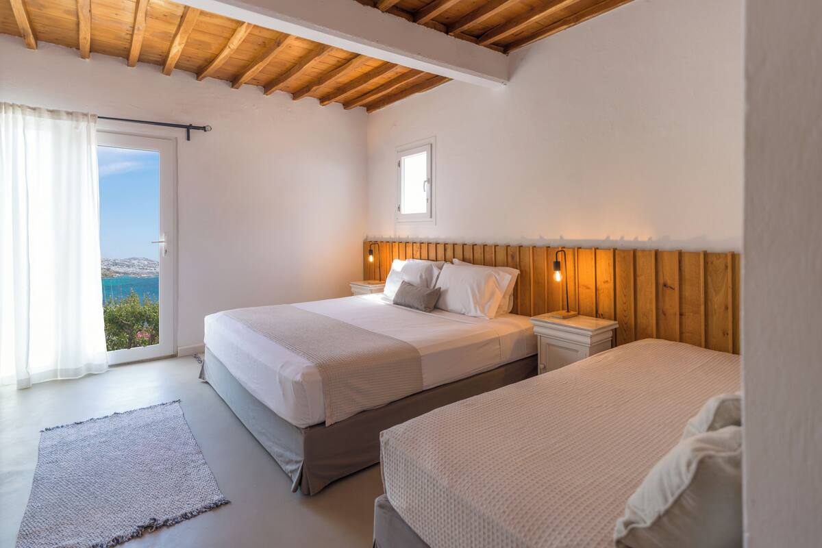 Quarto com duas camas, vigas de madeira no teto, janela com vista e mesa de cabeceira com abajur.