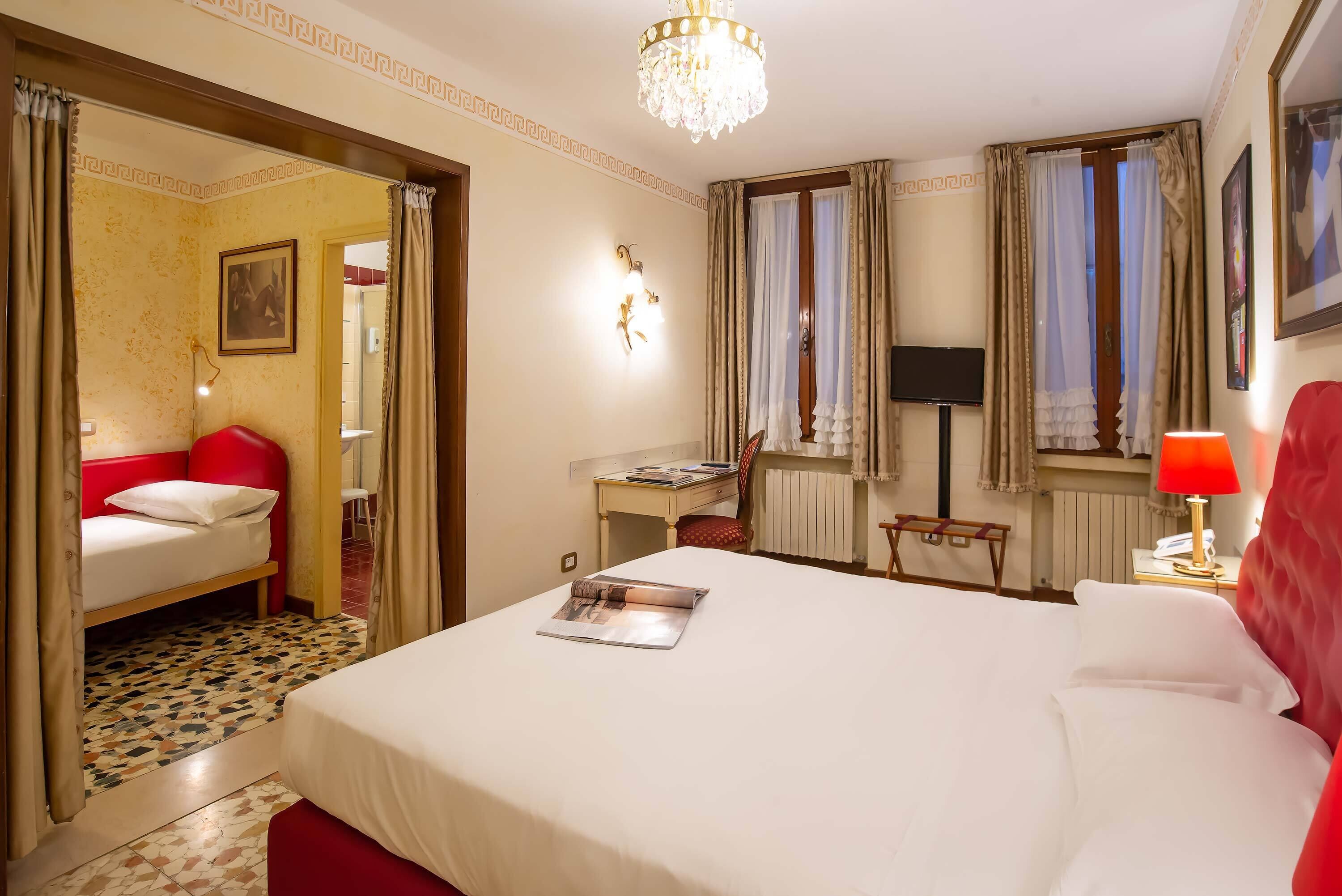 Quarto de hotel com cama, escrivaninha, poltrona, sofá vermelho, televisão e lustre.