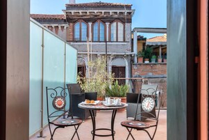 Um pátio com uma mesa redonda e quatro cadeiras, cercado por plantas em vasos e um edifício com janelas em arco.