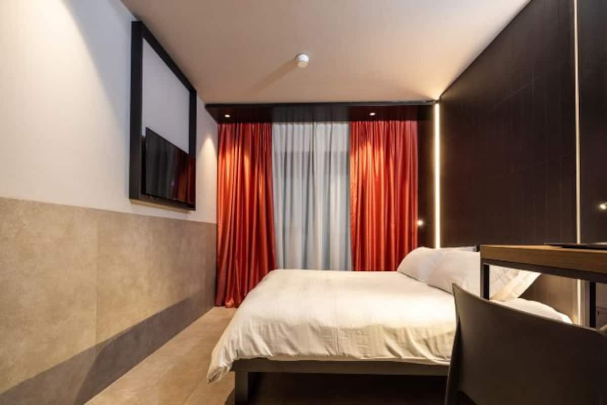 Quarto de hotel moderno com uma cama, um espelho e uma janela com cortinas vermelhas.