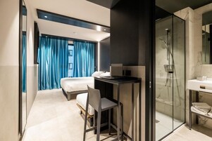 Quarto de hotel moderno com cama, escrivaninha, cadeira, chuveiro e banheiro.