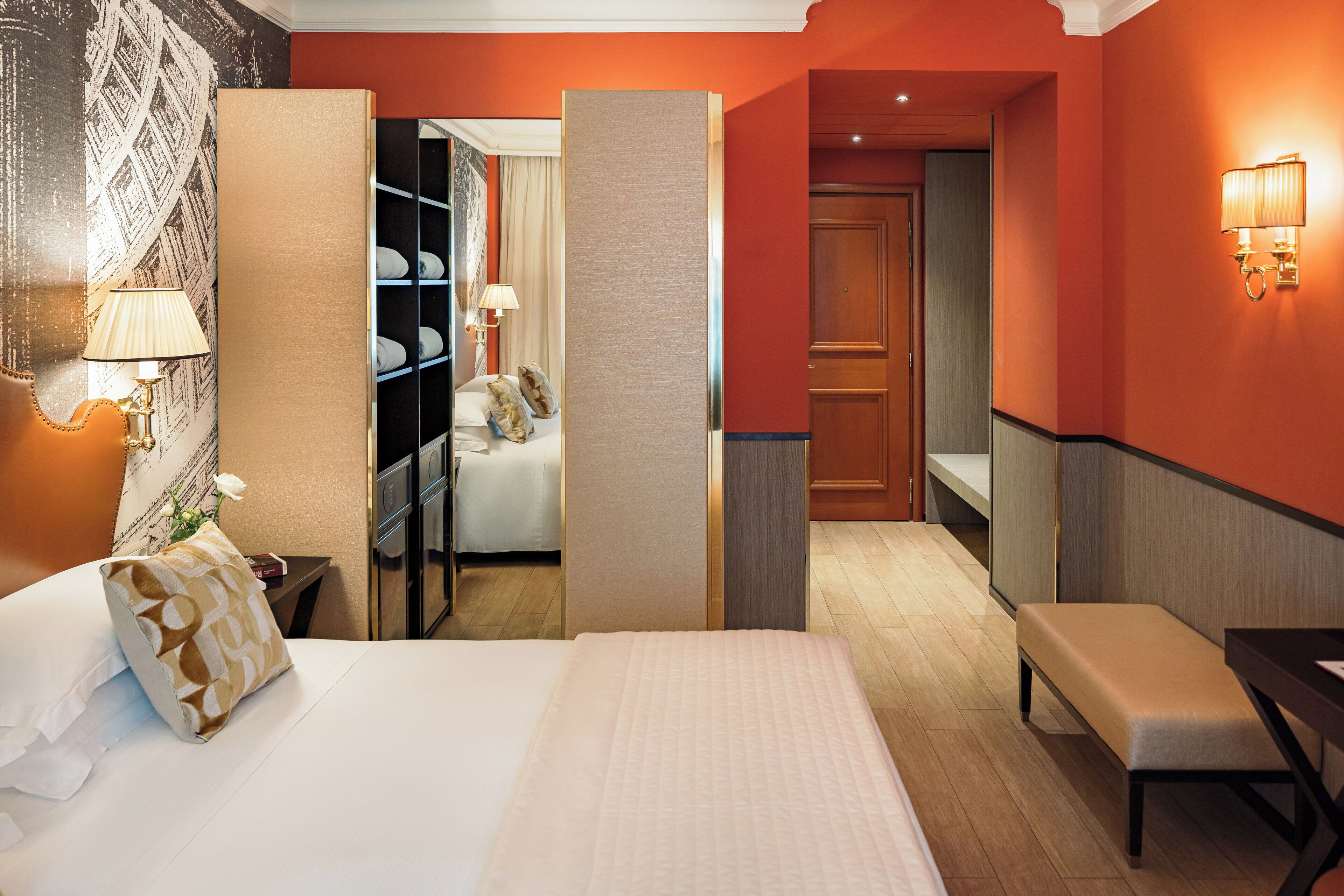 Quarto de hotel com cama, mesa de cabeceira, luminária e um banquinho.