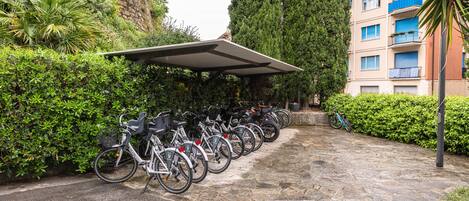 Várias bicicletas estão estacionadas sob uma cobertura, rodeadas por vegetação e um edifício com varandas.