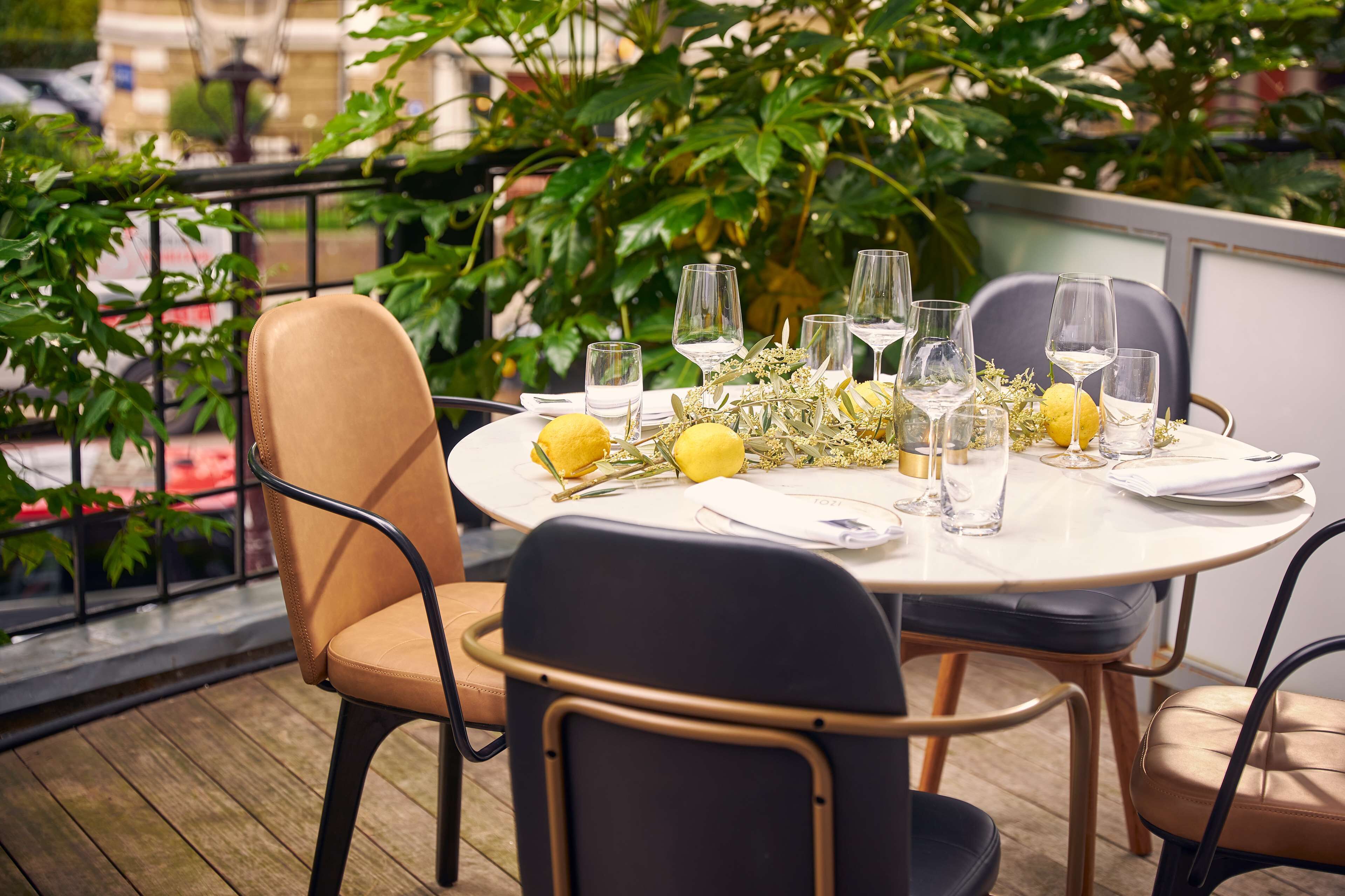 Mesa redonda posta com cadeiras, copos e limões em terraço ao ar livre com plantas e guarda-corpo.