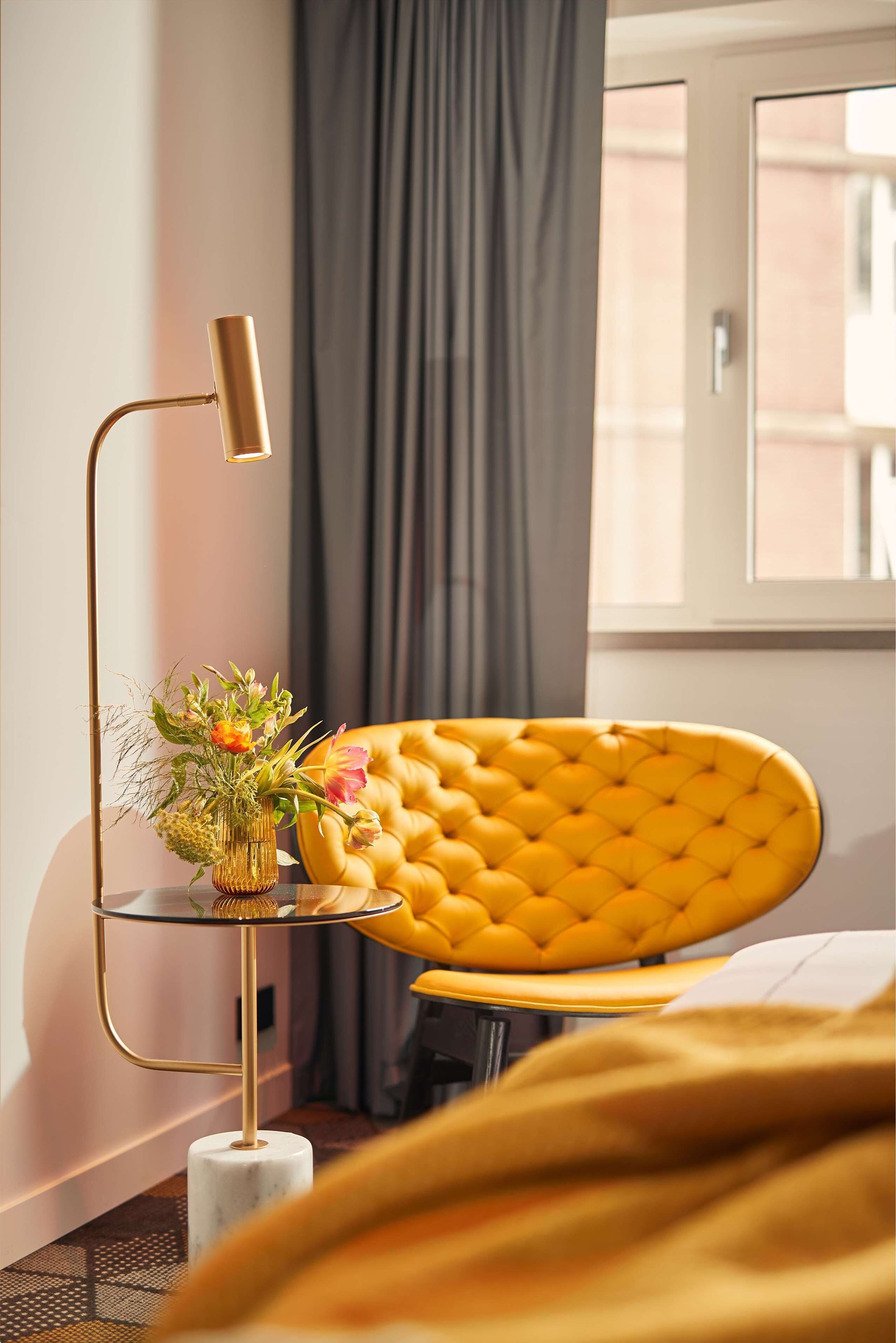 Um quarto com uma poltrona amarela estofada, uma pequena mesa redonda com um vaso de flores e um abajur.