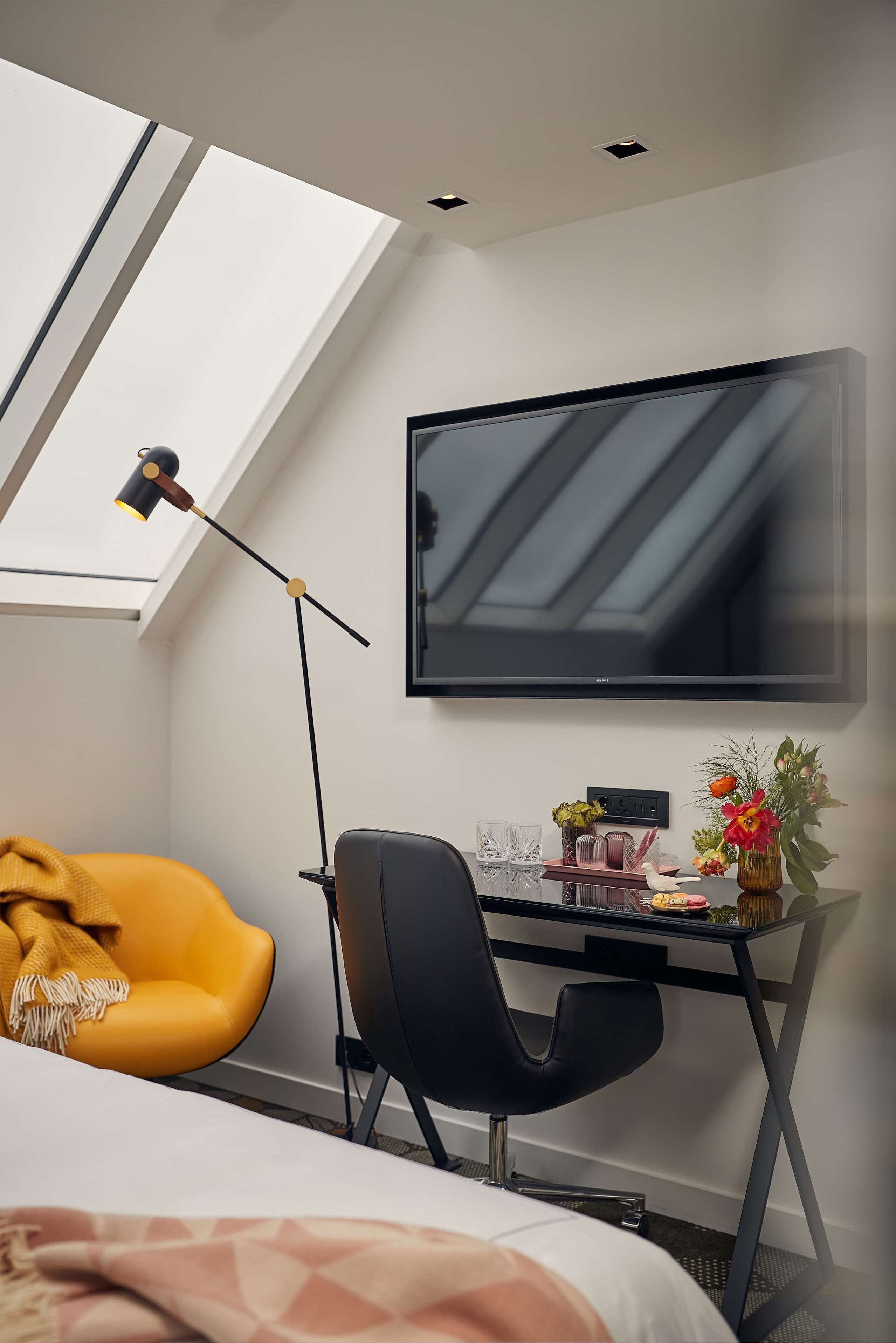 Quarto de hotel moderno com clarabóia, uma grande televisão de tela plana, uma escrivaninha com cadeira e uma poltrona amarela.