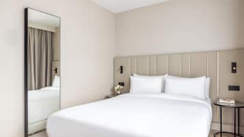 Quarto de hotel moderno com uma cama grande, uma mesinha de cabeceira com um vaso de flores, uma mesa lateral redonda e um espelho grande.