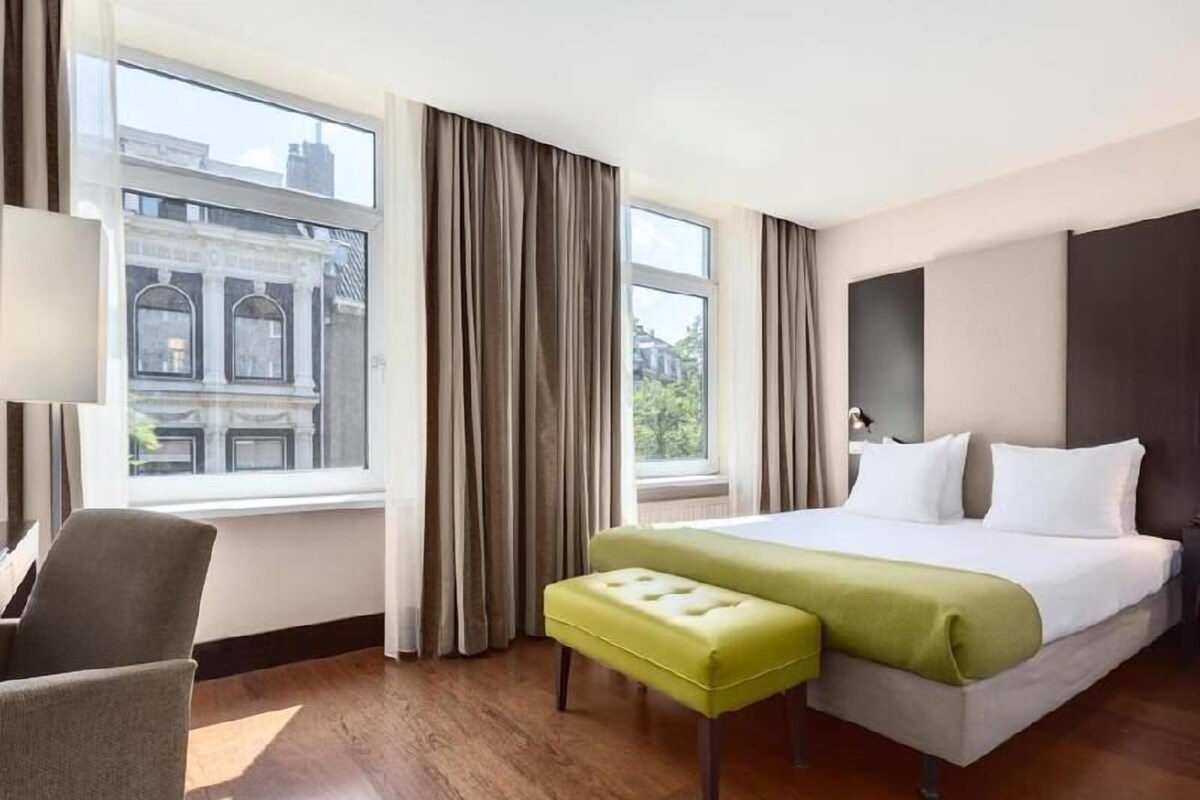 Quarto de hotel com cama, escrivaninha, cadeira, janela com cortinas e vista para edifícios.