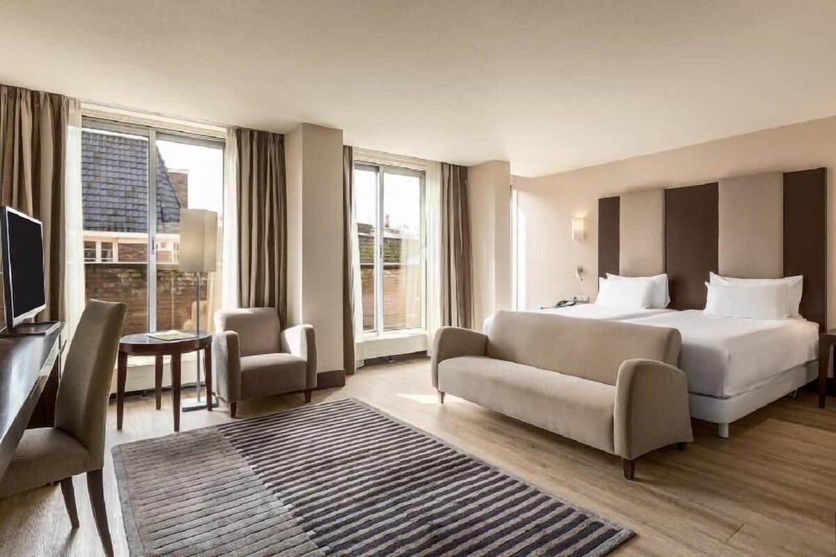 Quarto de hotel com uma cama grande, um sofá, uma escrivaninha com cadeira, uma TV e uma janela com cortinas.