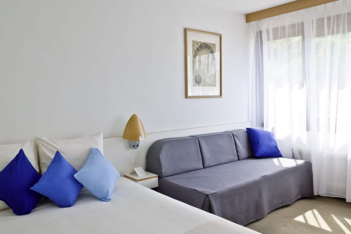 Quarto de hotel com cama, sofá cinzento e mesa de cabeceira com abajur.