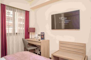 Quarto de hotel com uma cama, uma escrivaninha com cadeira, uma televisão na parede e uma janela com cortinas.