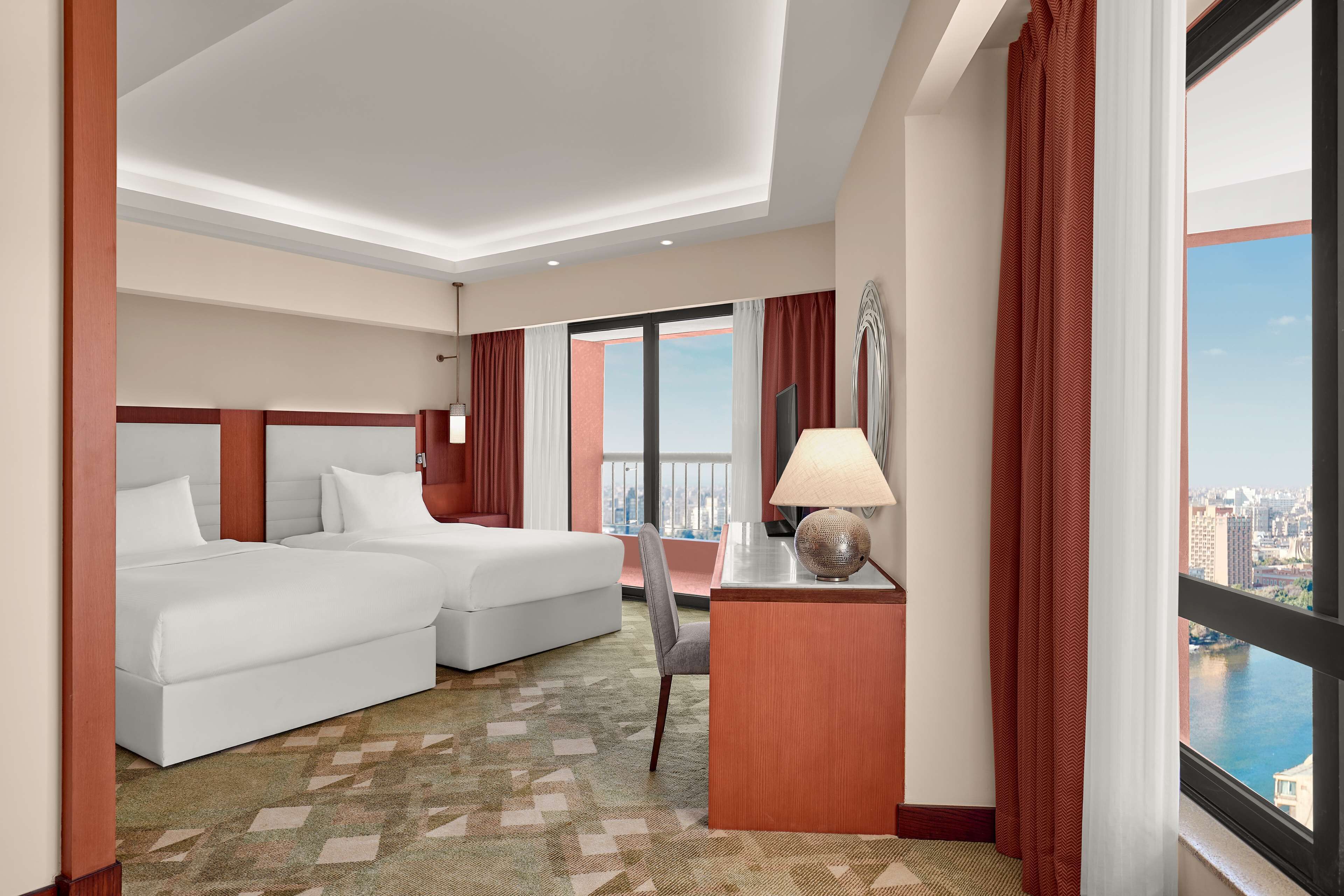 Quarto de hotel com duas camas, uma escrivaninha, uma cadeira e uma janela grande com vista para a cidade.