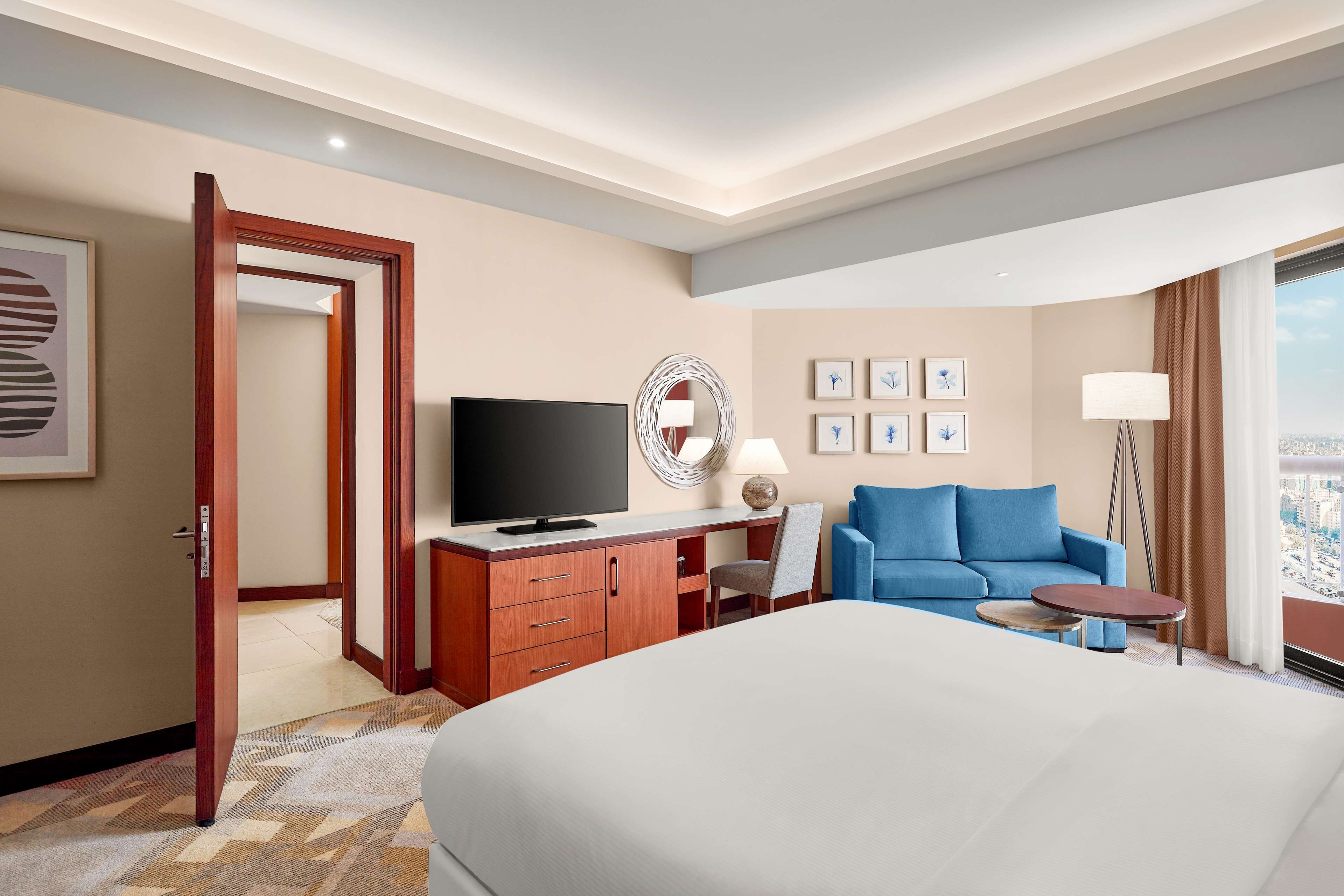 Quarto de hotel com uma cama grande, uma escrivaninha com televisão, um sofá azul, uma mesa redonda pequena e vista para a cidade.