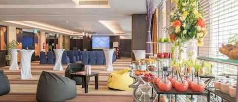 Um saguão de hotel com áreas de estar, um buffet com diversas opções de comida e uma tela de televisão ao fundo.