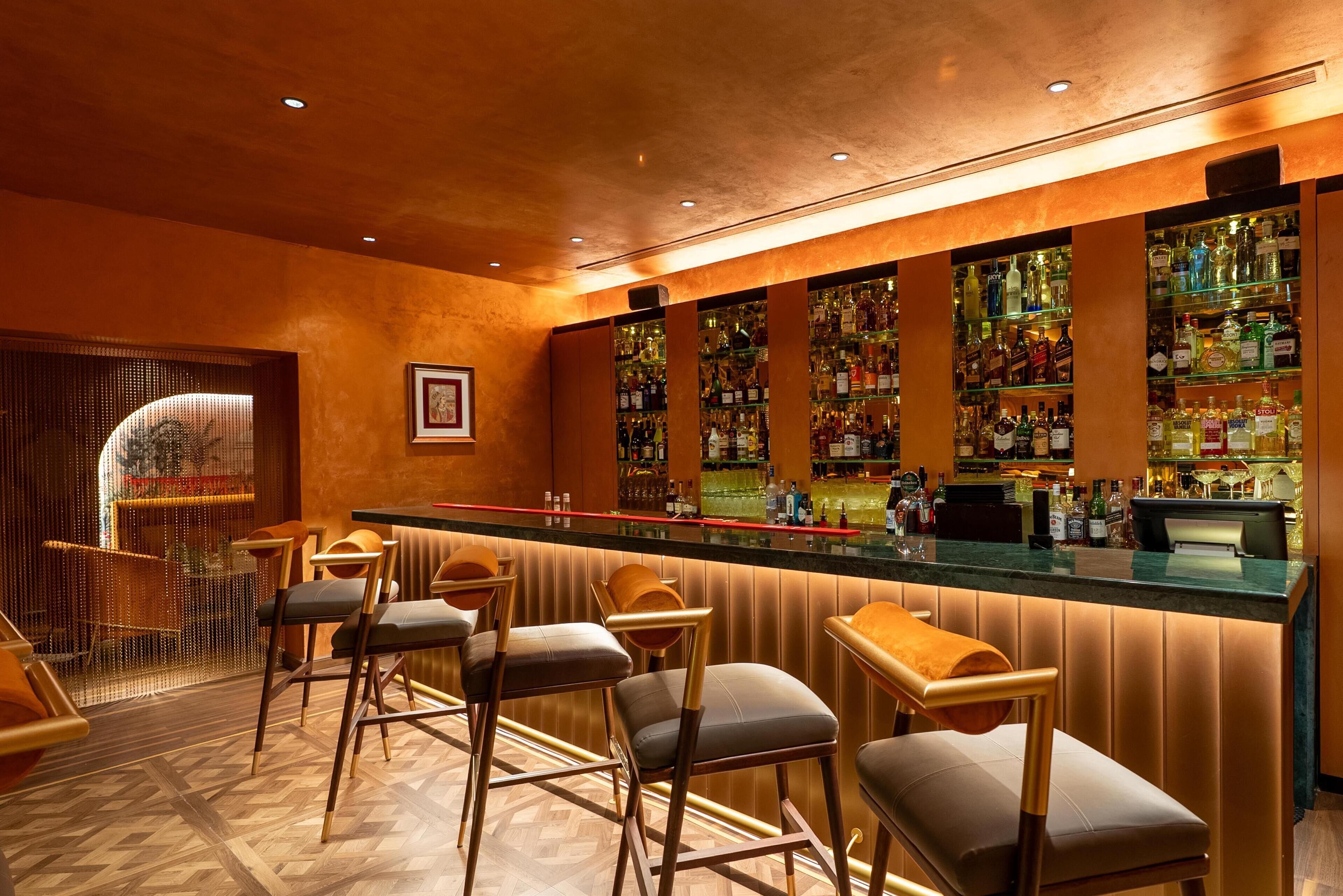 Uma área de bar com banquetas de madeira e diversas garrafas de bebidas alcoólicas nas prateleiras.