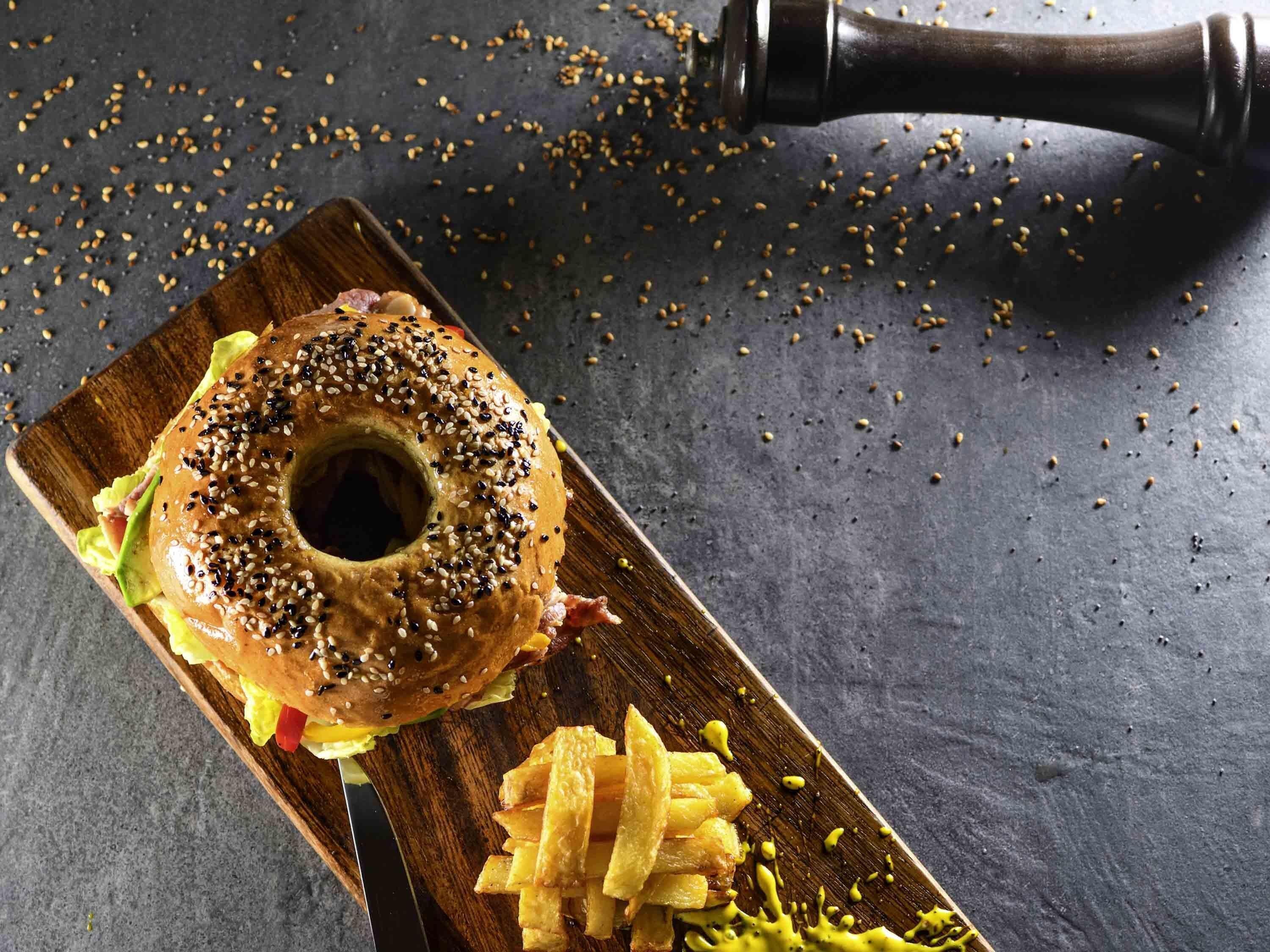 Um bagel com sementes de gergelim sobre uma tábua de madeira, acompanhado de batatas rústicas e um moedor de pimenta.