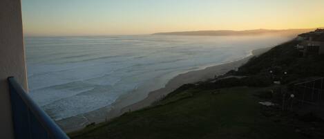 Vista costeira de uma varanda com vista para o oceano, área gramada e edifícios à direita.