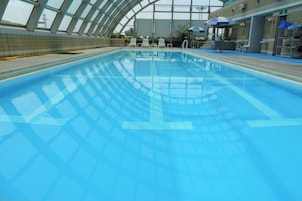 Uma grande piscina interior com água azul cristalina, rodeada por um revestimento de azulejos e um teto de vidro.