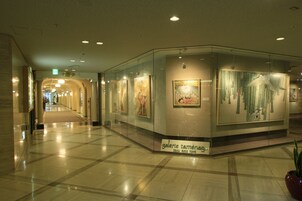 Corredor moderno de hotel com paredes de vidro exibindo obras de arte, uma placa de saída visível e um chão de mármore polido.