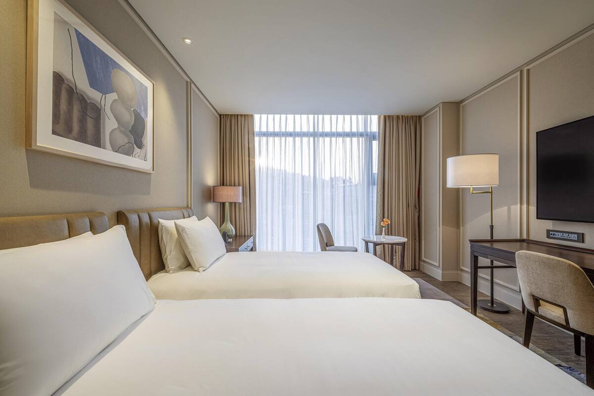 Quarto de hotel com duas camas, uma escrivaninha, uma televisão e um quadro na parede.