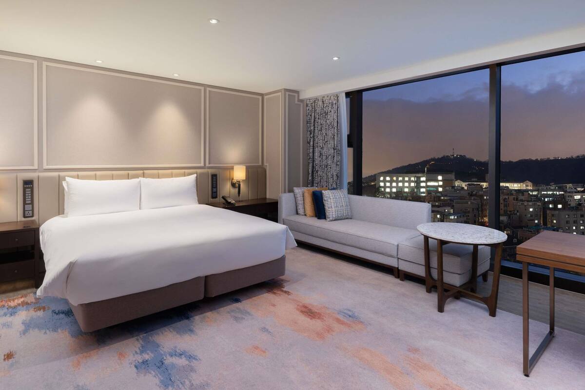 Quarto de hotel com uma cama, um sofá, uma mesinha e uma janela grande com vista para a cidade.