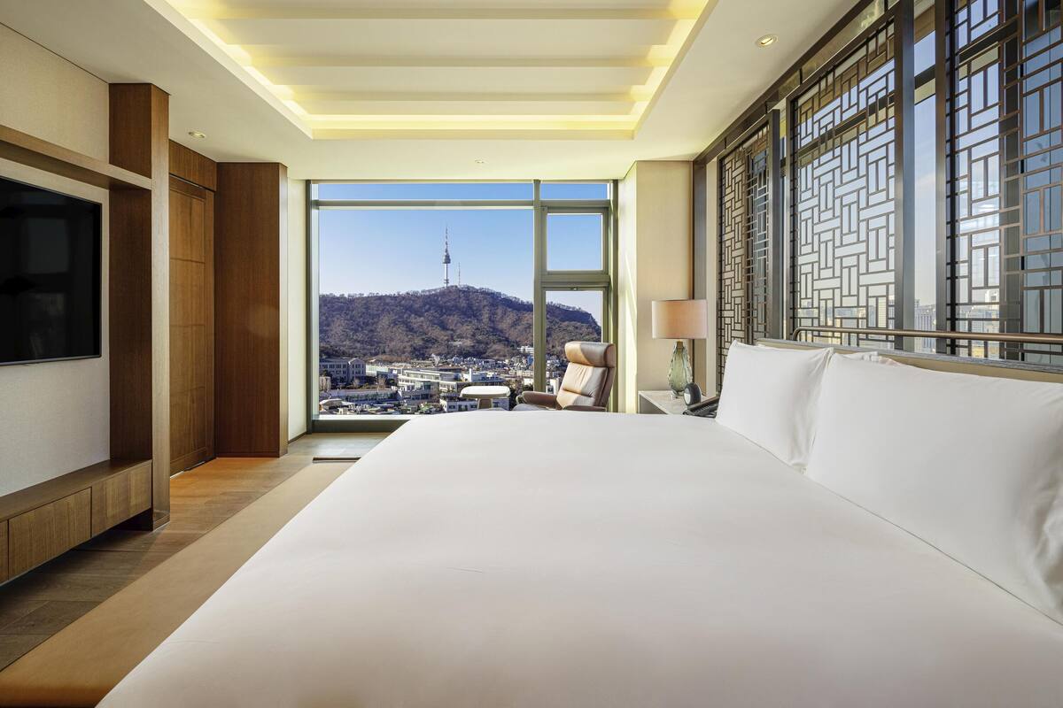 Quarto de hotel com uma cama grande, televisão, vista para a paisagem urbana e uma colina.