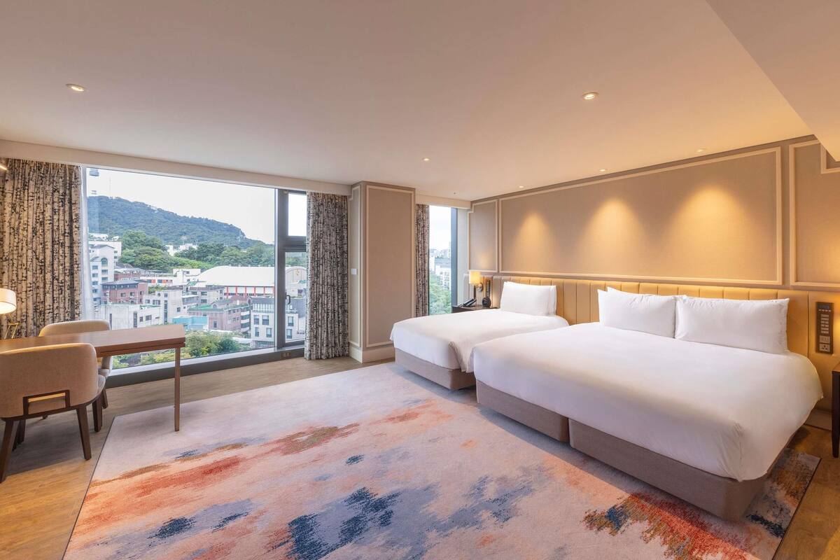 Quarto de hotel com duas camas, uma escrivaninha, uma cadeira e uma janela ampla com vista para a cidade.