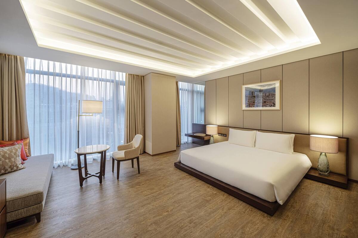 Quarto de hotel com uma cama grande, uma mesa pequena, uma cadeira, um sofá e uma pintura urbana na parede.
