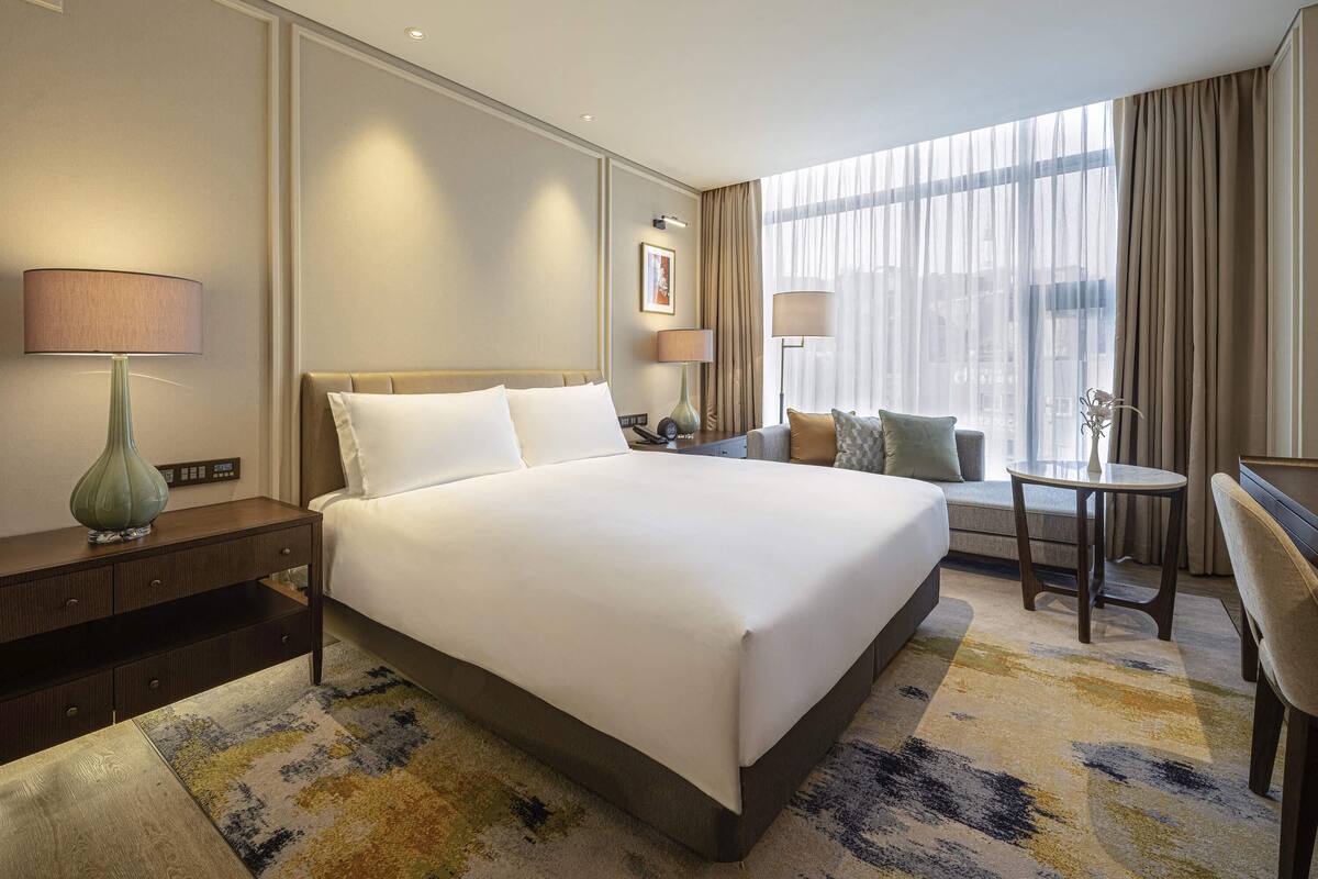 Quarto de hotel com uma cama grande, luminárias laterais, uma mesinha e vista para a cidade através de cortinas finas.