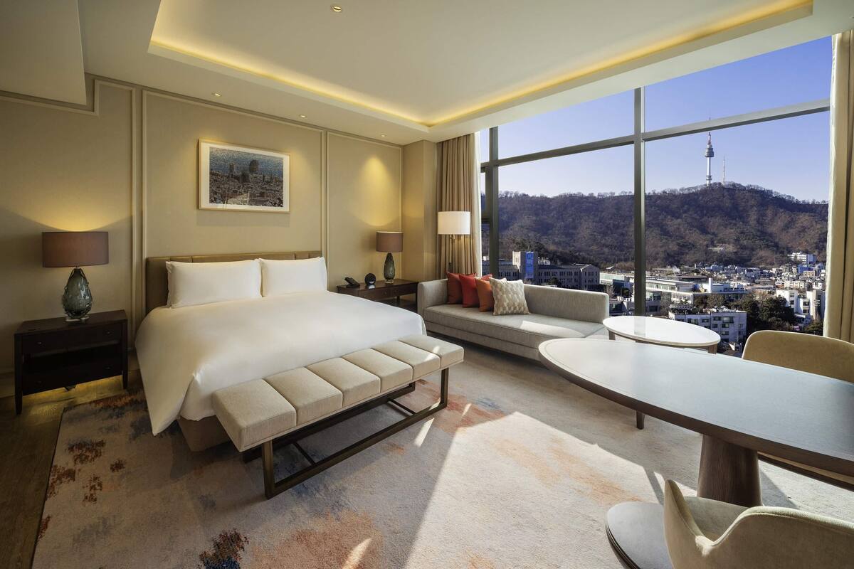 Quarto de hotel com cama, sofá, mesa de jantar e vista para a cidade e montanhas.