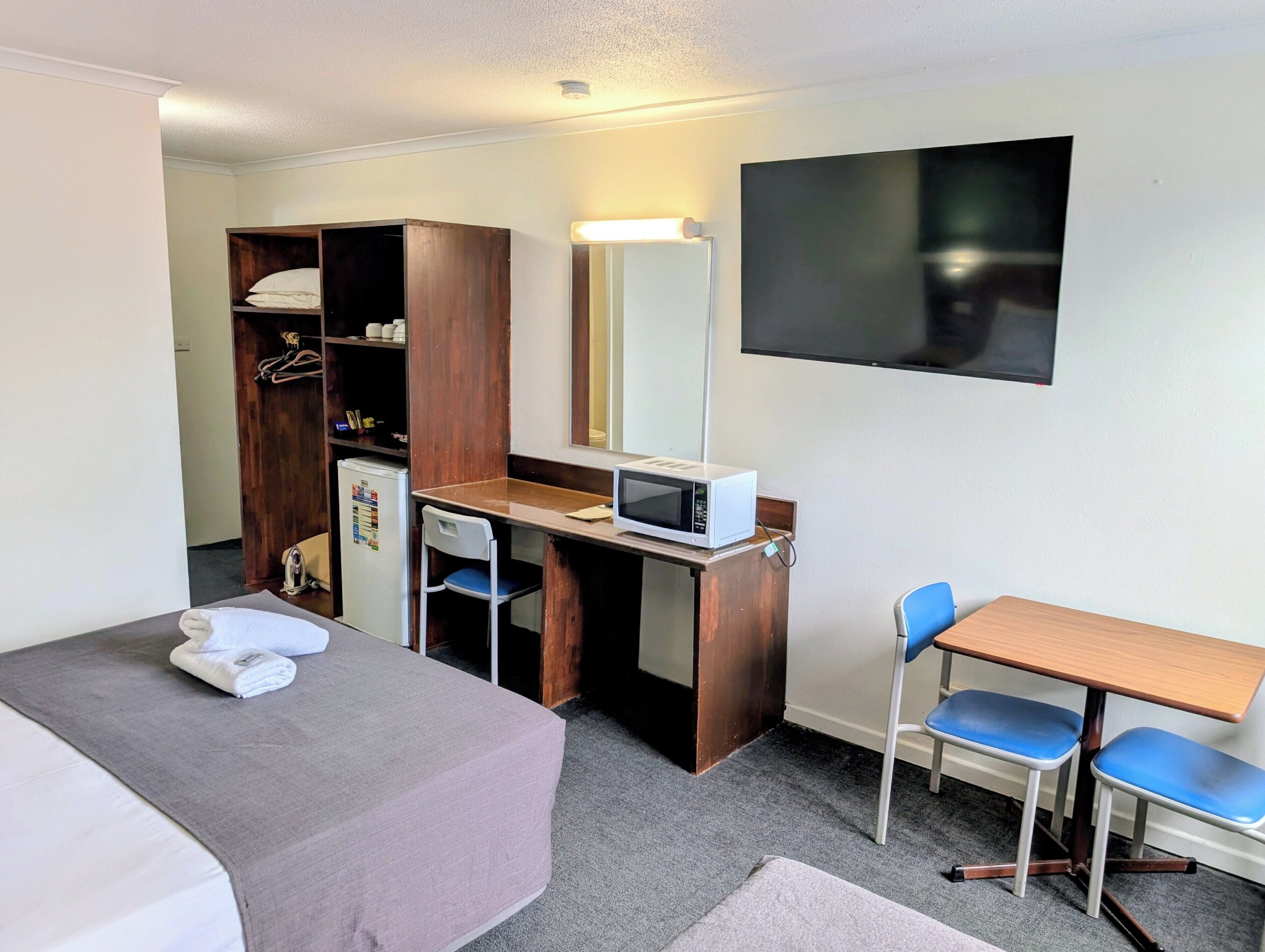 Quarto de hotel compacto com uma cama, escrivaninha com micro-ondas, cadeira, guarda-roupa e um espelho grande.