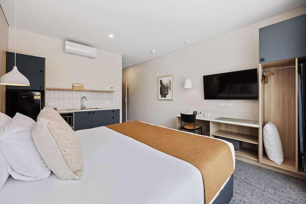Quarto de hotel com cama, secretária, televisão, cozinha compacta e guarda-roupa.