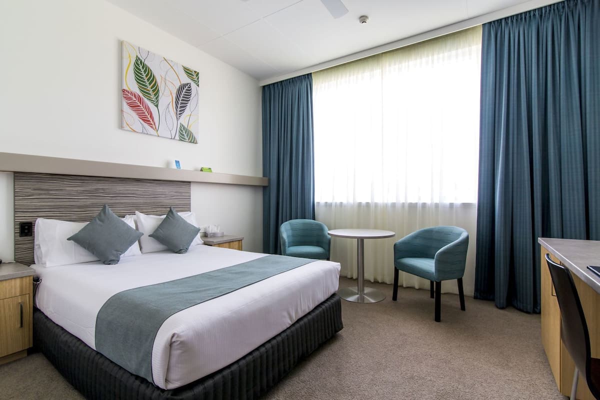 Quarto de hotel com cama, duas cadeiras, uma mesinha, cabeceira e janela com cortinas.