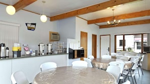 Uma área de refeições com mesas redondas cobertas com plástico, cadeiras brancas, um balcão de cozinha com eletrodomésticos e uma placa com a inscrição "BEAR BIKES".