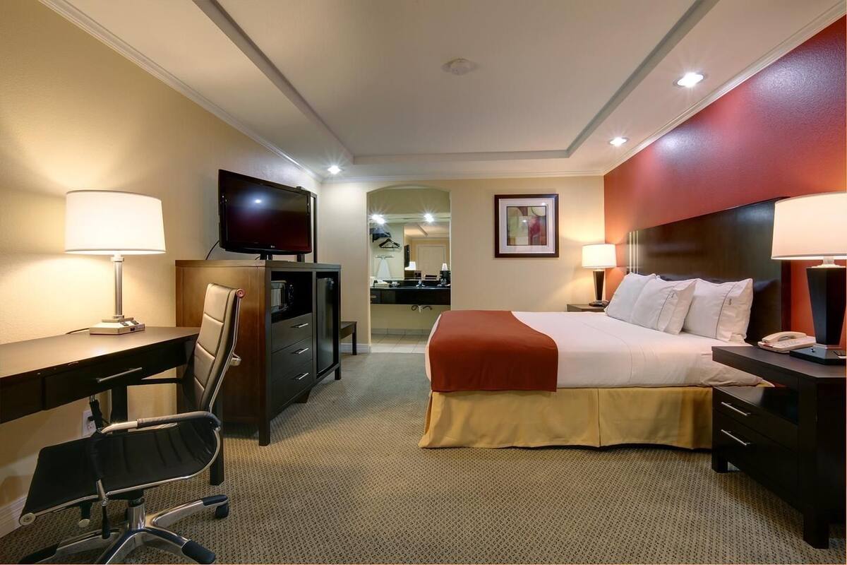 Quarto de hotel com cama, escrivaninha, cadeira, televisão e banheiro ao fundo.