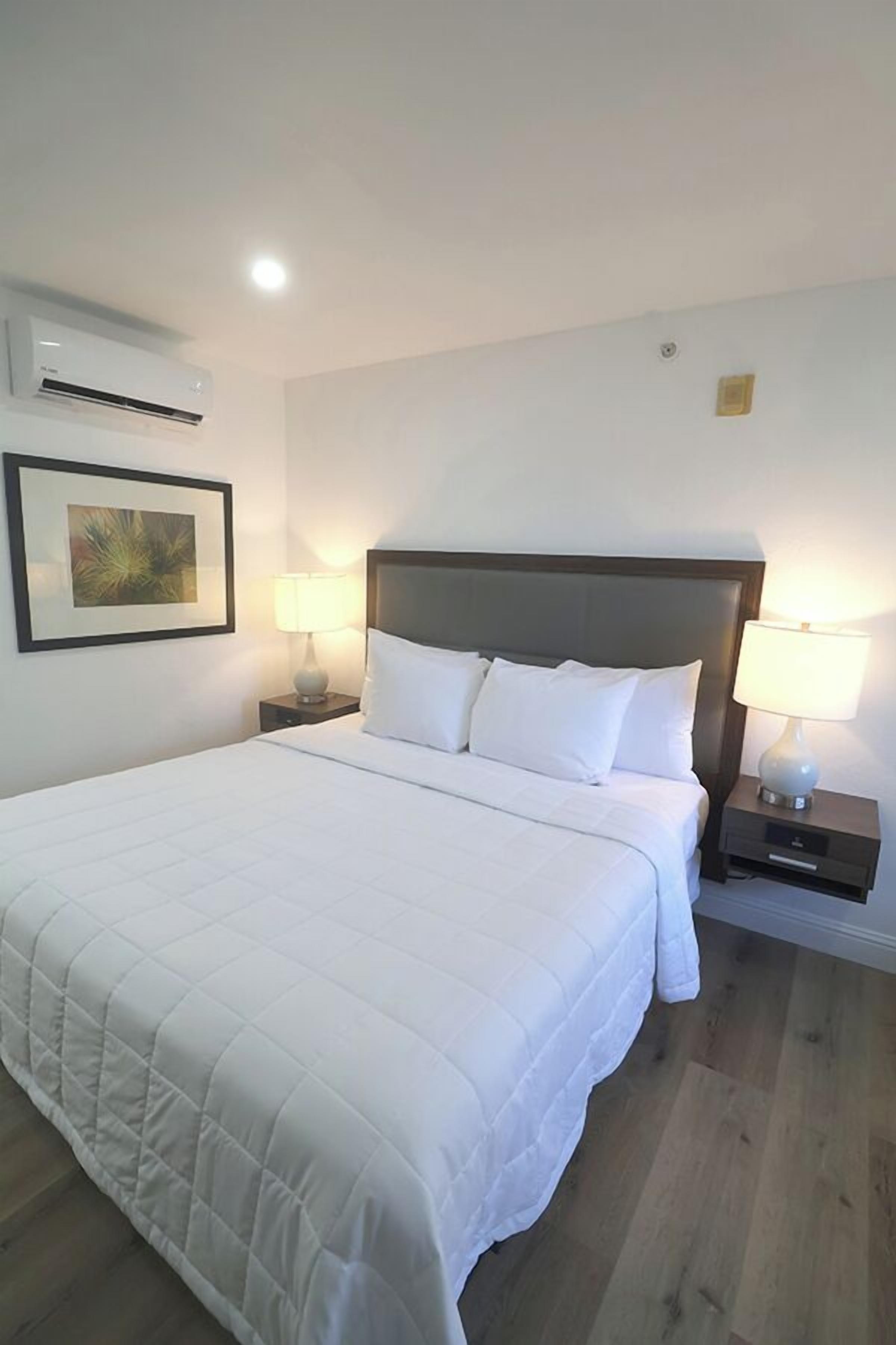 Quarto de hotel com uma cama grande, duas mesas de cabeceira com luminárias, ar condicionado fixado na parede e um quadro emoldurado.