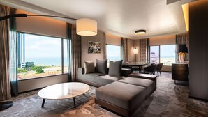 Quarto de hotel moderno com uma janela ampla que oferece vista para a praia e o oceano, um sofá, uma mesa de jantar e uma pequena mesa de centro.