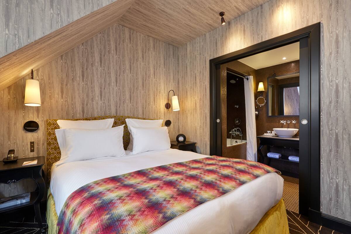 Quarto de hotel com uma cama grande, mesa de cabeceira, escrivaninha, espelho e banheiro com pia e espelho.