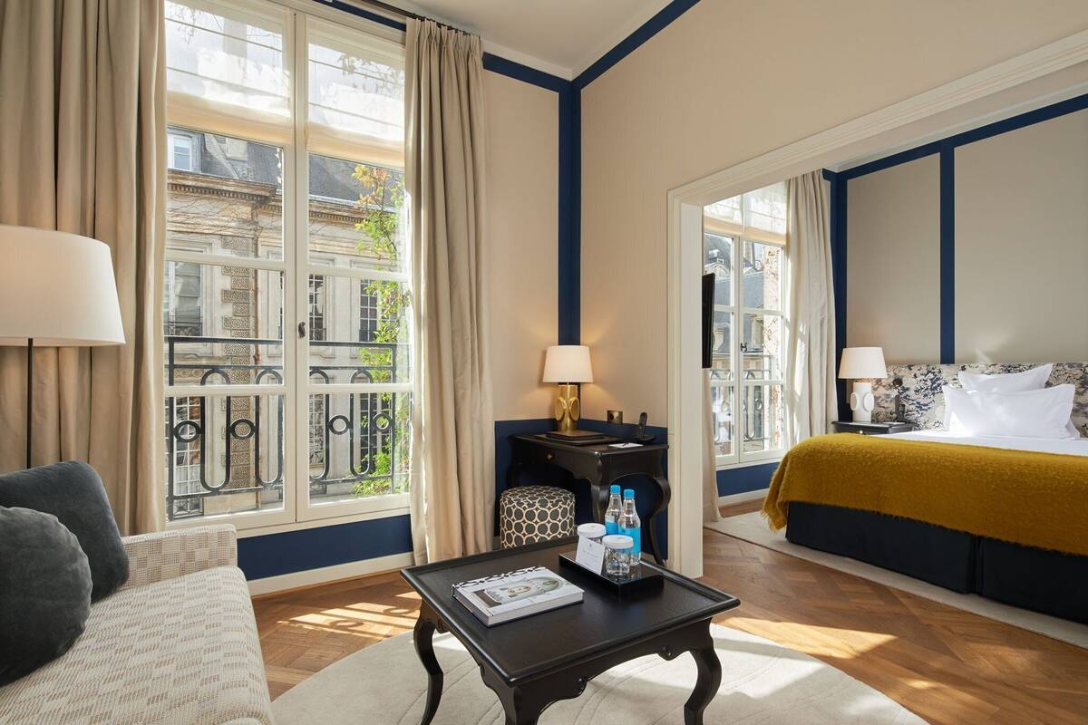 Quarto de hotel com cama, sofá, mesa com uma revista, luminária e janela com cortinas.