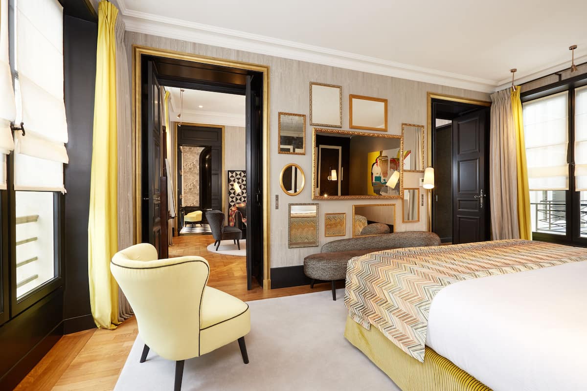 Quarto de hotel com uma cama grande, um espaço de estar com uma cadeira, uma penteadeira com espelhos e vista para o corredor.