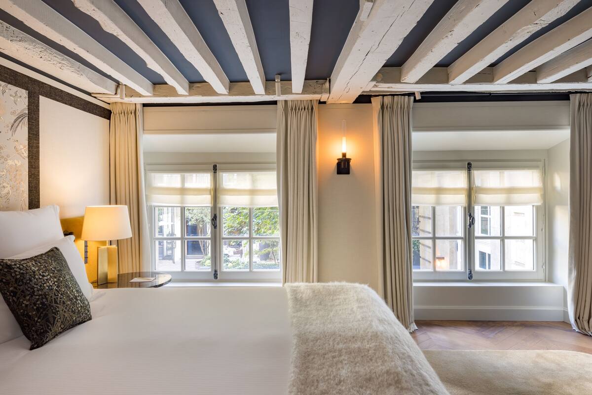Quarto de hotel com uma cama grande, mesa de cabeceira com abajur, janela com cortinas e vista para o exterior.
