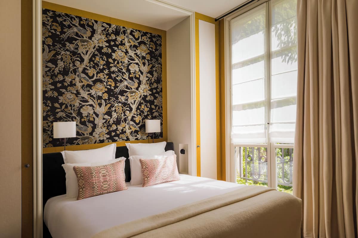 Quarto de hotel com uma cama grande, duas luminárias de cabeceira, papel de parede floral e uma janela com cortinas.