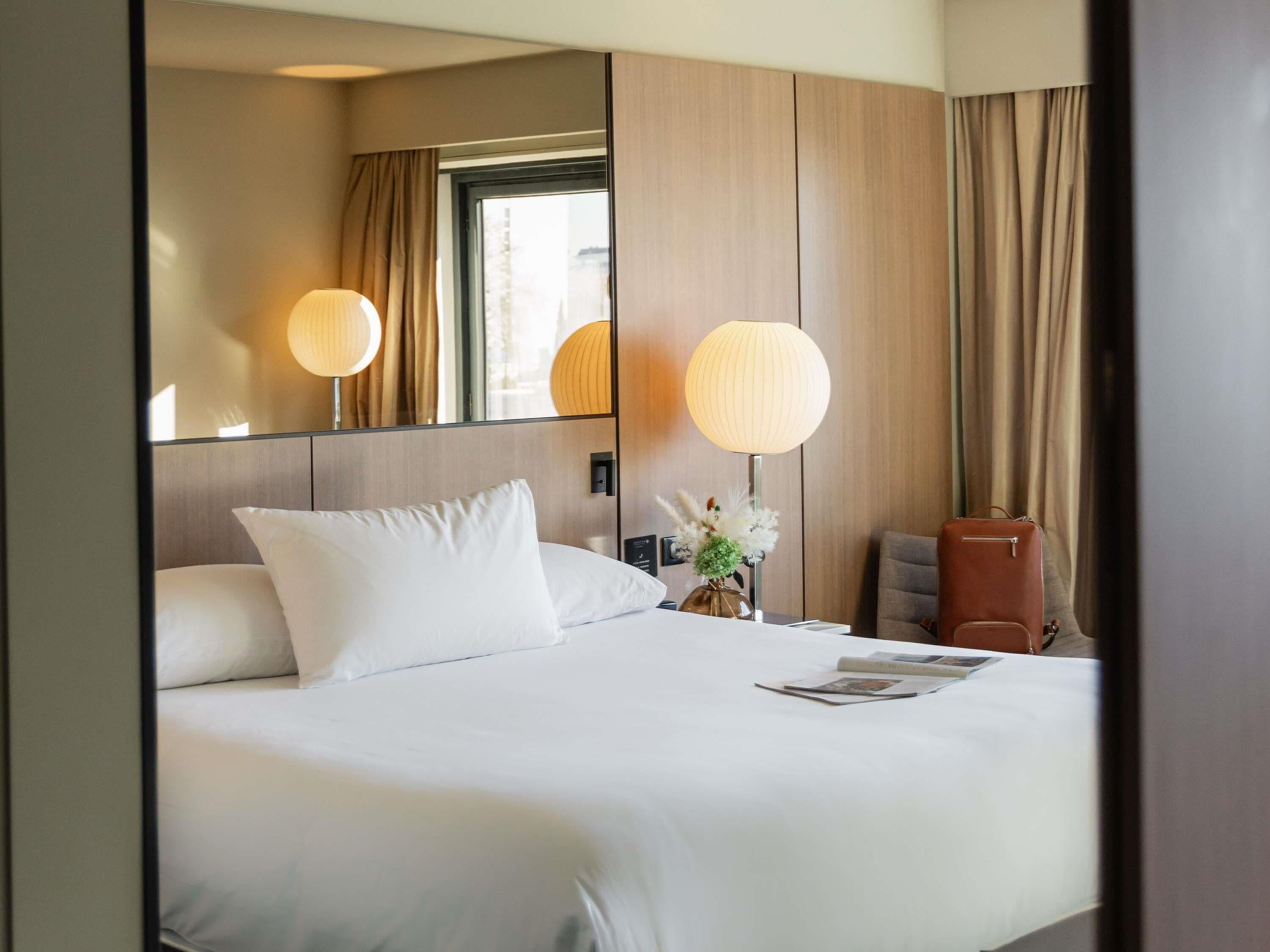 Quarto de hotel com uma cama, mesa de cabeceira, luminária, espelho e janela com cortinas.