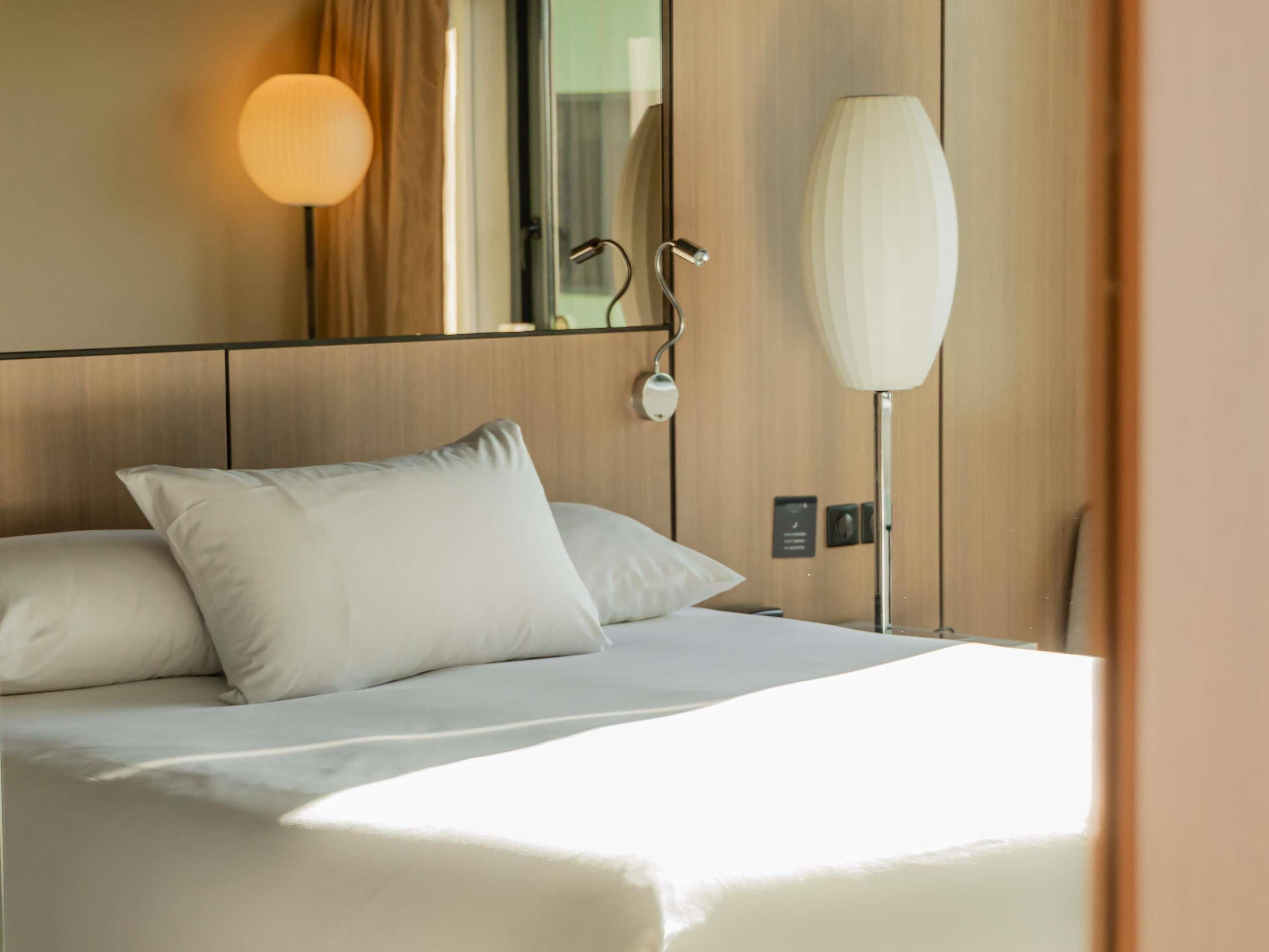 Quarto de hotel com uma cama, mesa de cabeceira, luminária e espelho.
