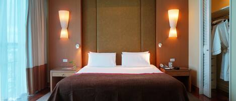 Quarto de hotel com uma cama grande, duas mesas de cabeceira, um criado-mudo, um espelho, uma janela com cortinas e um guarda-roupa.