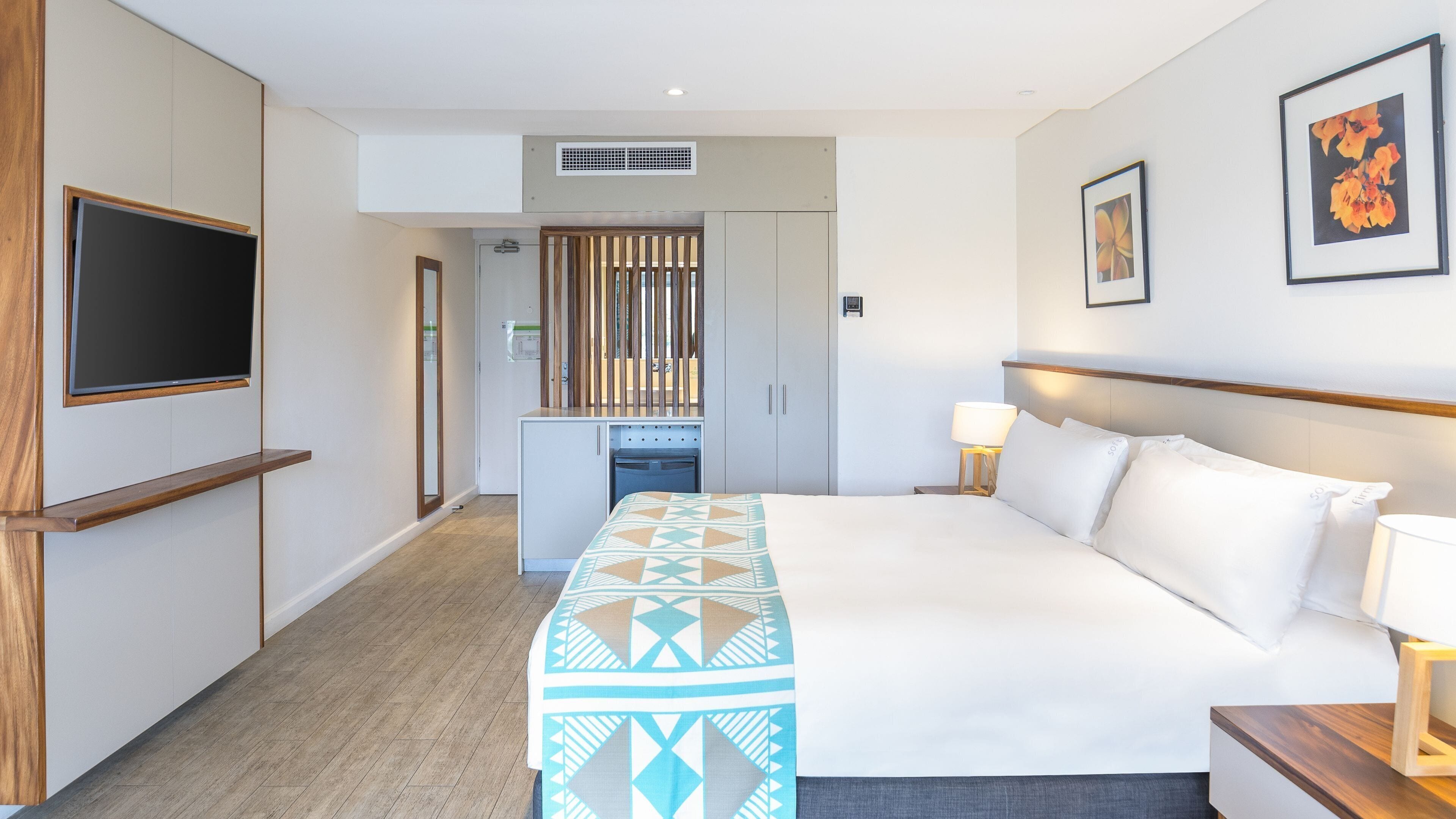 Quarto de hotel com cama, televisão, escrivaninha e cozinha compacta.
