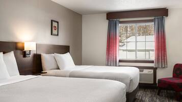 Quarto de hotel com duas camas, uma mesa de cabeceira, luminária, uma cadeira e janela com cortinas.