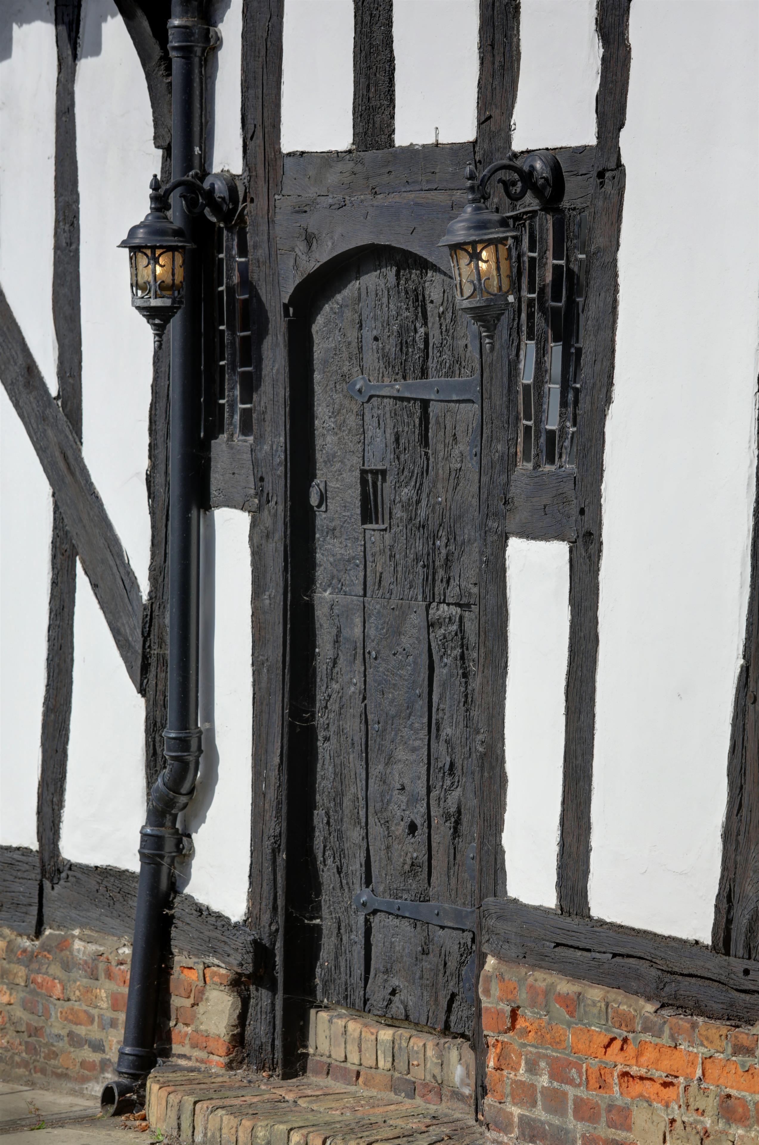 Uma porta de madeira preta, parede branca, base de tijolos e duas luzes em estilo lanterna, também pretas.